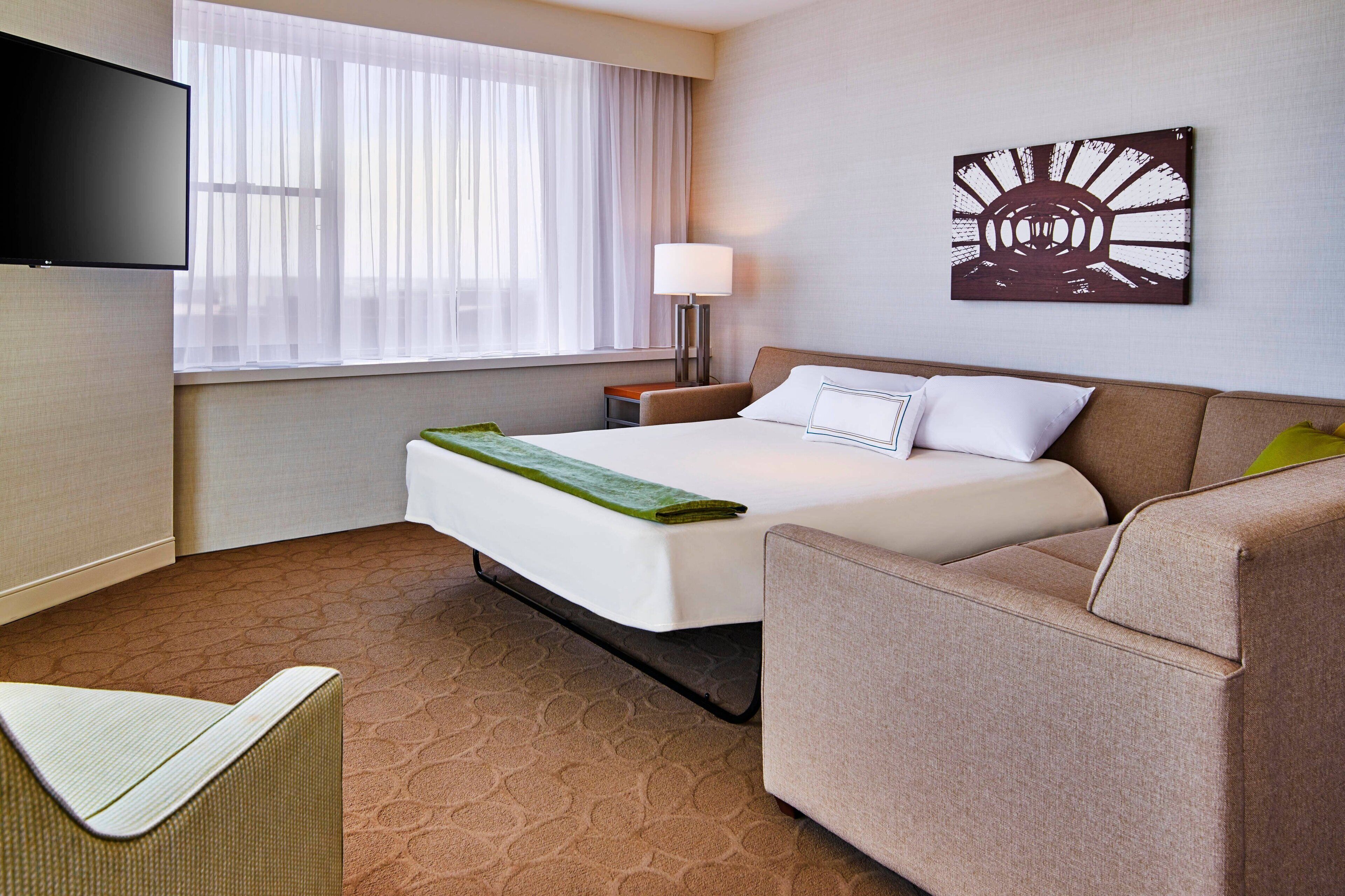 Quarto de hotel com cama, sofá, televisão, luminária e janela com cortinas leves.