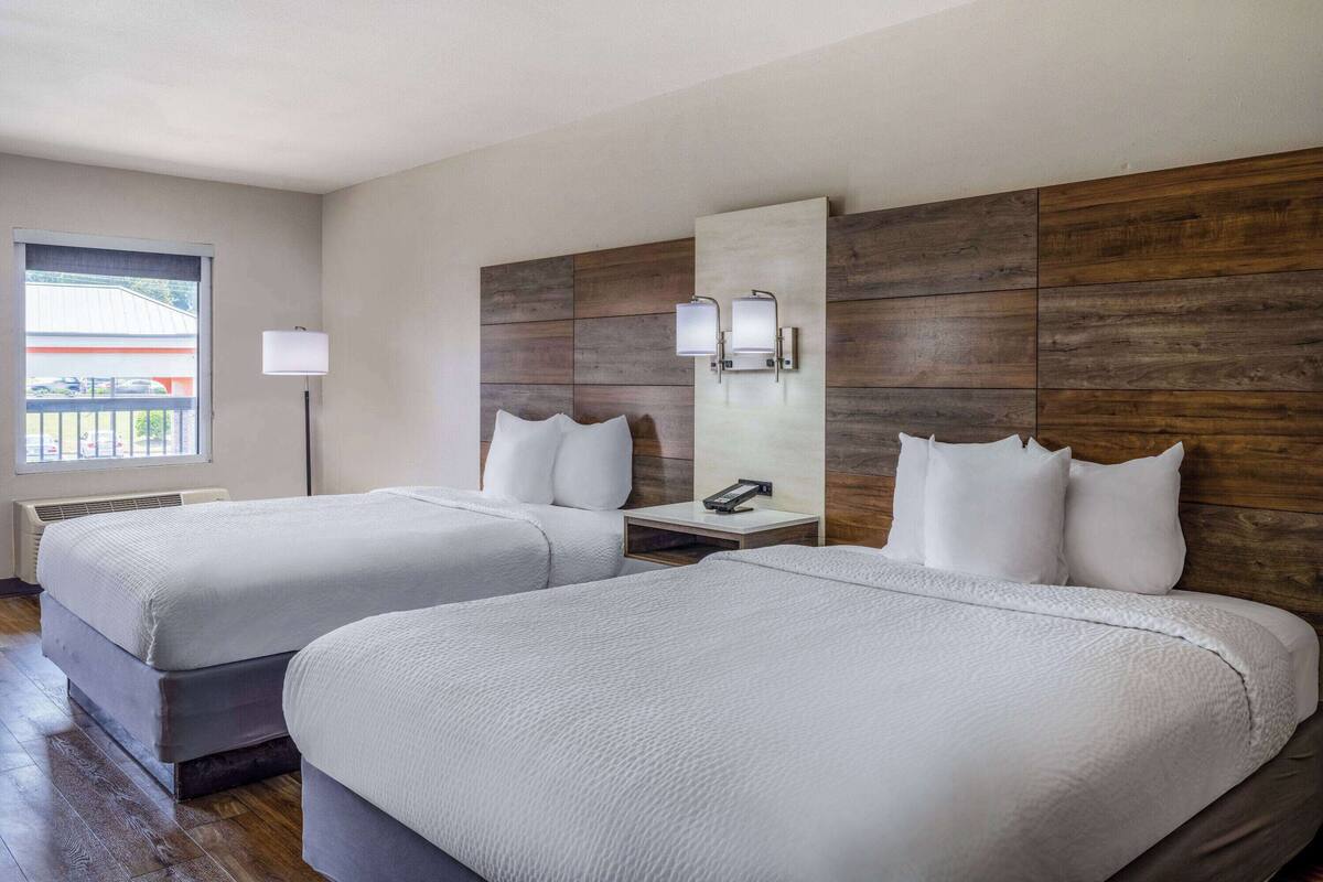 Quarto de hotel com duas camas, uma mesa de cabeceira, uma luminária e uma janela com vista para um edifício.
