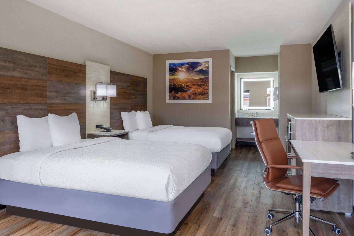 Quarto de hotel com duas camas, uma escrivaninha, uma cadeira, uma televisão e um quadro na parede.