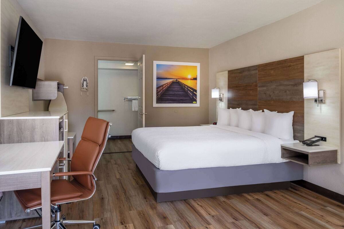 Quarto de hotel com uma cama, uma escrivaninha, uma cadeira, uma televisão e uma pintura de um pôr do sol.