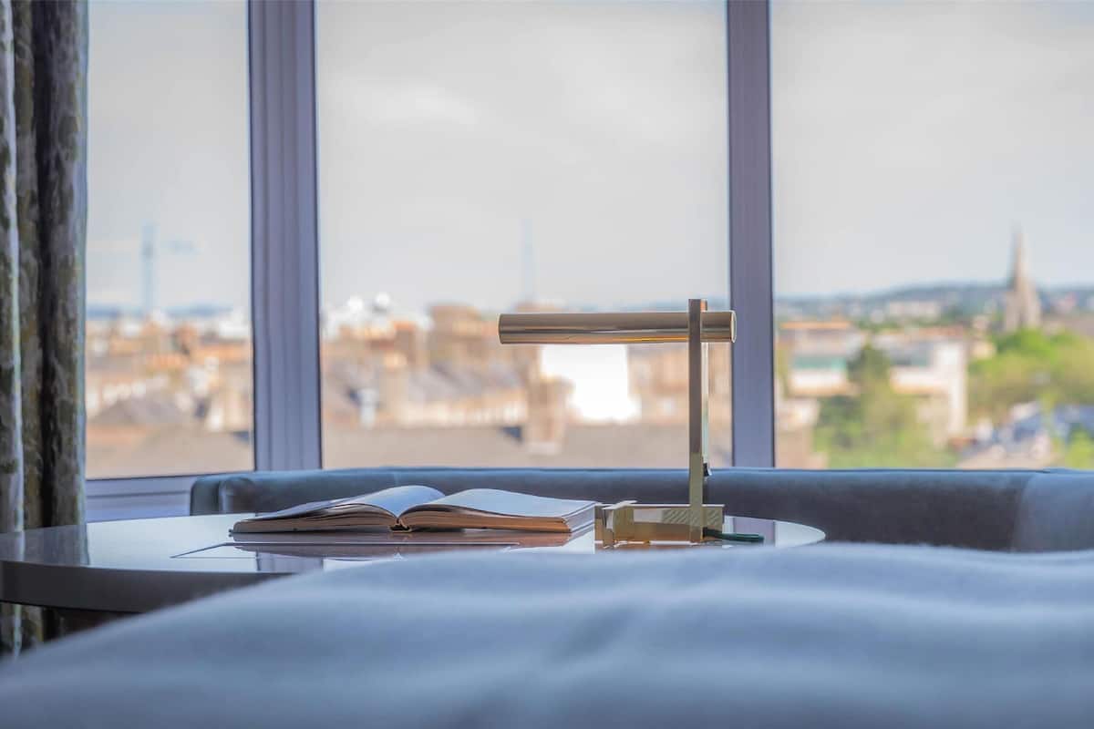 Quarto de hotel com uma janela grande, mesa de cabeceira com um livro e uma luminária, e vista para a paisagem urbana.