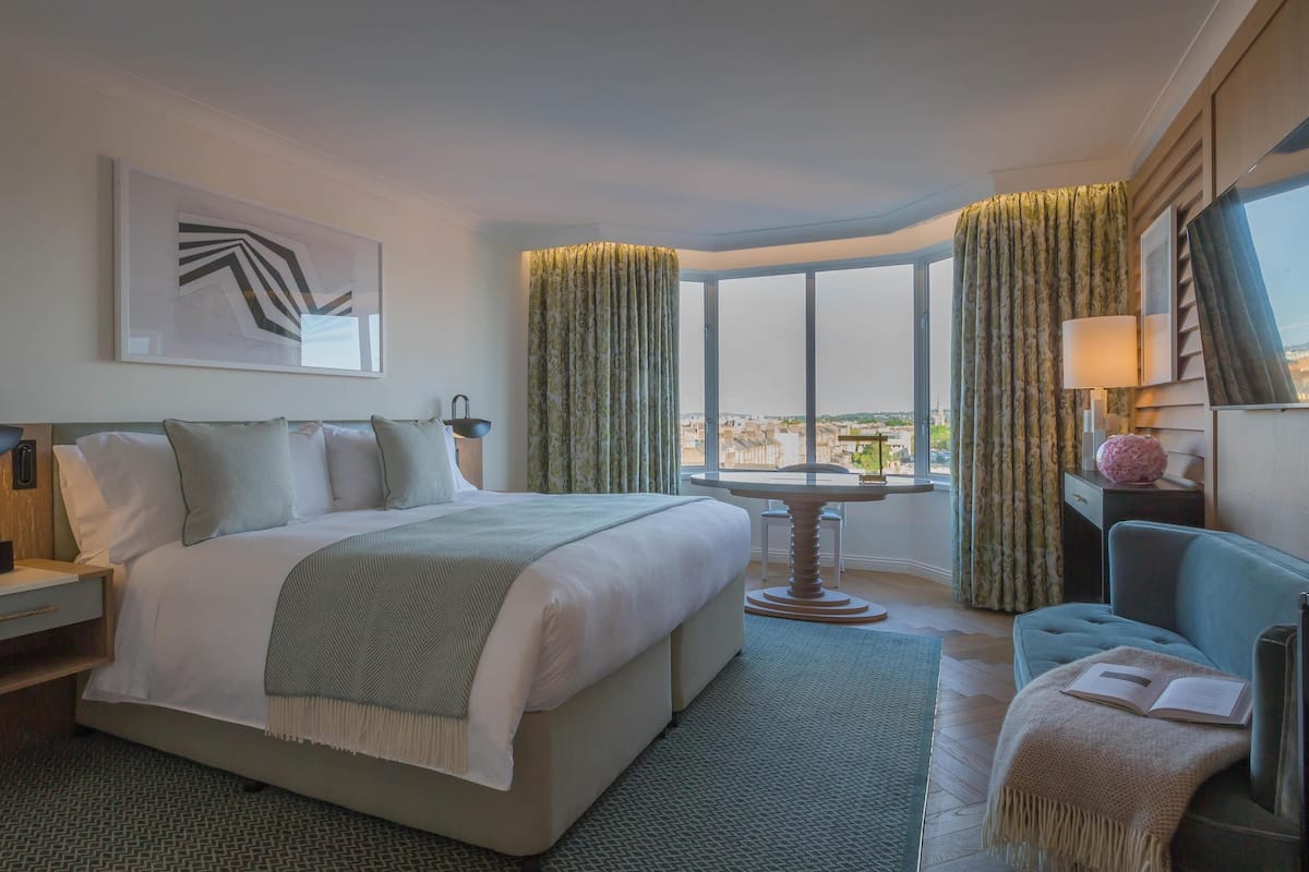 Quarto de hotel com uma cama grande, uma mesinha redonda, uma poltrona azul e vista da cidade através de grandes janelas.