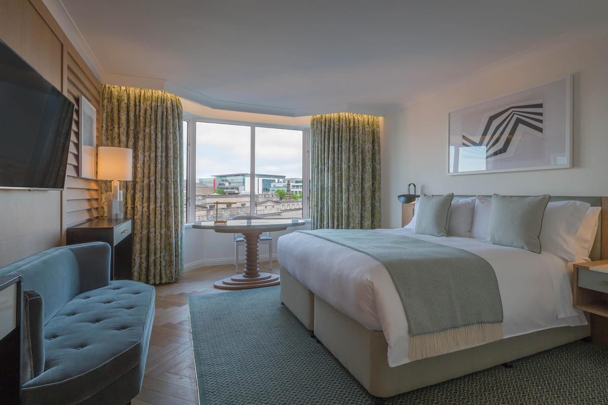 Quarto de hotel com uma cama grande, uma escrivaninha, uma cadeira, uma televisão e uma janela com cortinas.
