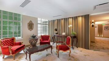 Um hall amplo com uma grande janela, confortáveis poltronas vermelhas revestidas, uma mesa de centro de madeira clássica e um espelho decorativo.
