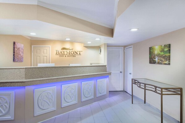 Uma área de recepção com o letreiro Baymont Inn & Suites, um balcão de atendimento, uma mesa com tampo de vidro e uma porta.