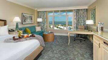 Quarto de hotel com cama, escrivaninha, vista para o oceano, janela com cortinas e um quadro na parede.