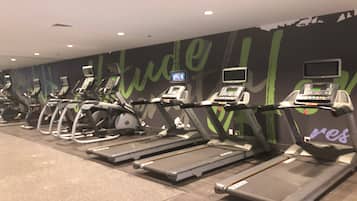 Academia com várias esteiras e bicicletas ergométricas, parede com mensagens motivacionais e tela digital exibindo conteúdo de treino.