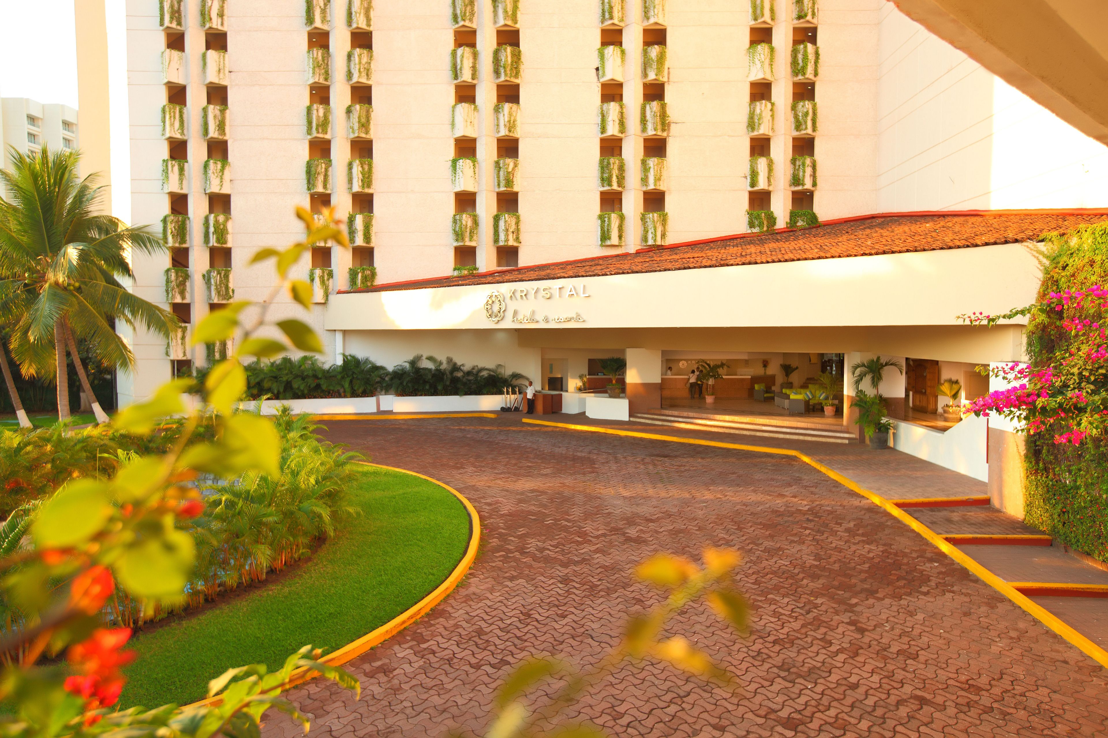 Entrada de hotel com caminho de tijolos, uma palmeira e um edifício com varandas.