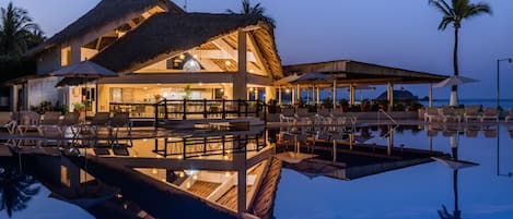 Um resort com piscina refletindo o edifício, cadeiras de sol e uma estrutura com telhado de palha.