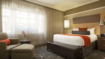 Quarto de hotel com cama, uma cadeira, uma mesinha com um vaso e uma luminária.