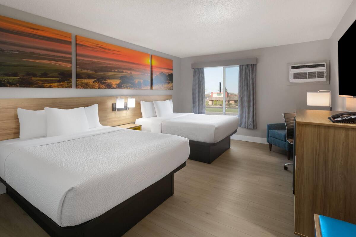 Quarto de hotel com duas camas, uma escrivaninha, uma cadeira, uma televisão e uma janela grande com vista.