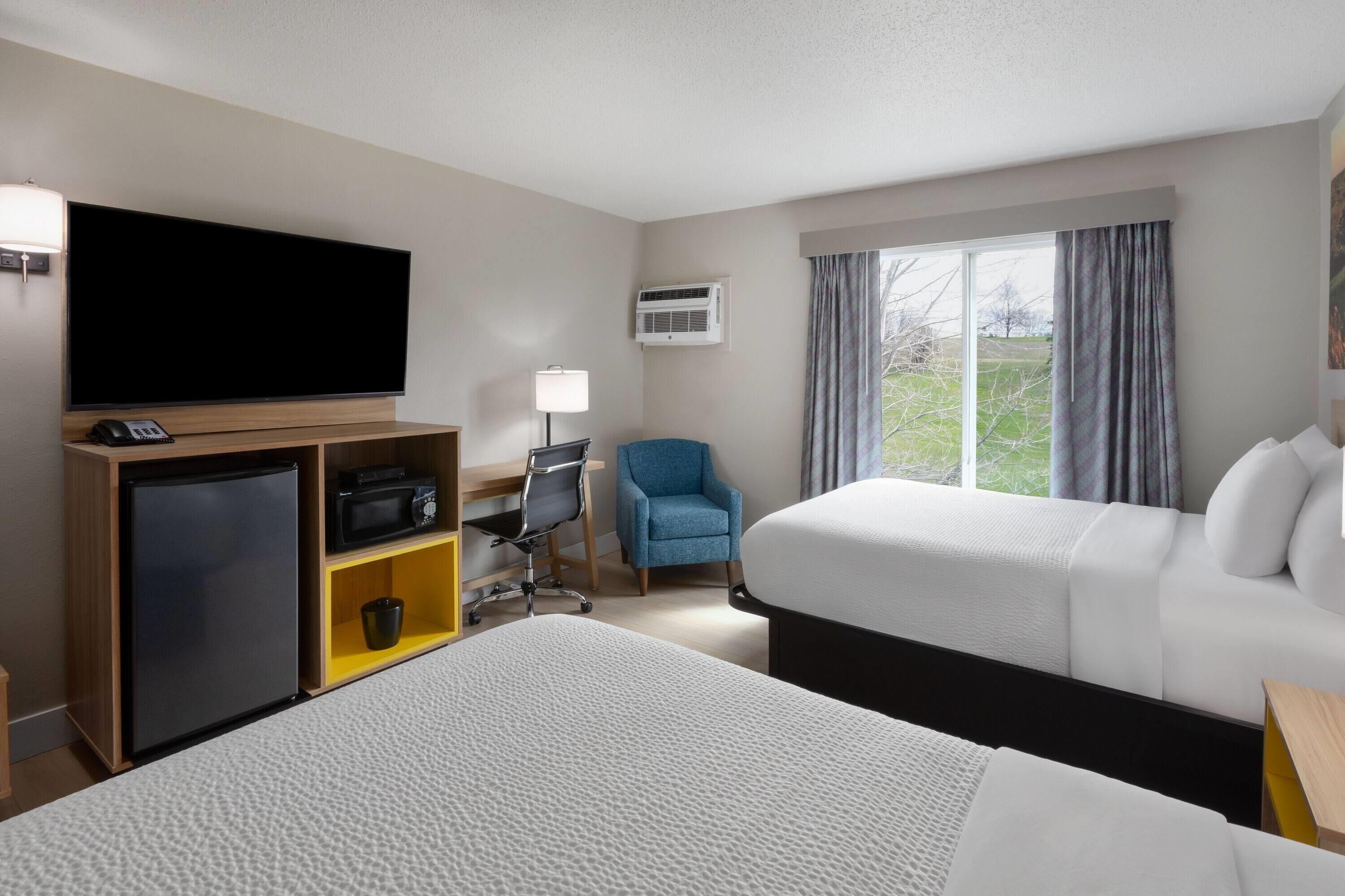 Quarto de hotel com cama, escrivaninha com computador, televisão e janela com cortinas.