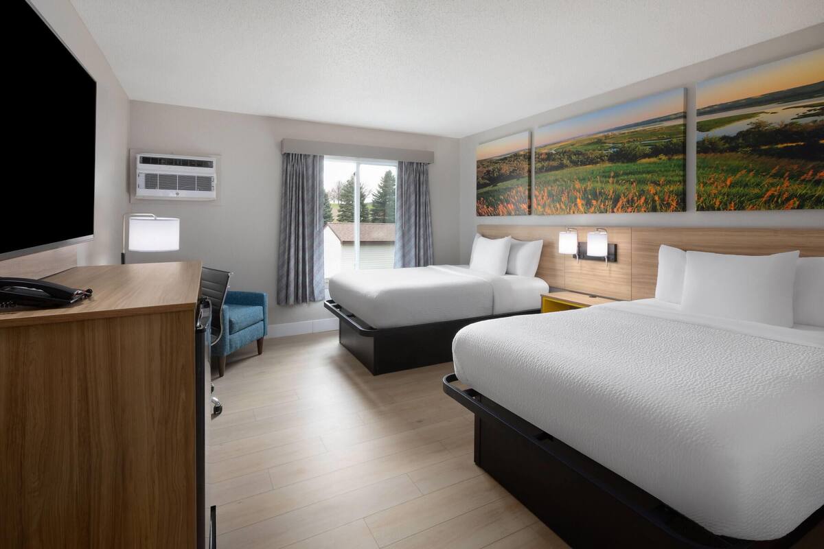 Quarto de hotel com cama, escrivaninha, cadeira, televisão, ar condicionado, janela com cortinas e um quadro na parede.