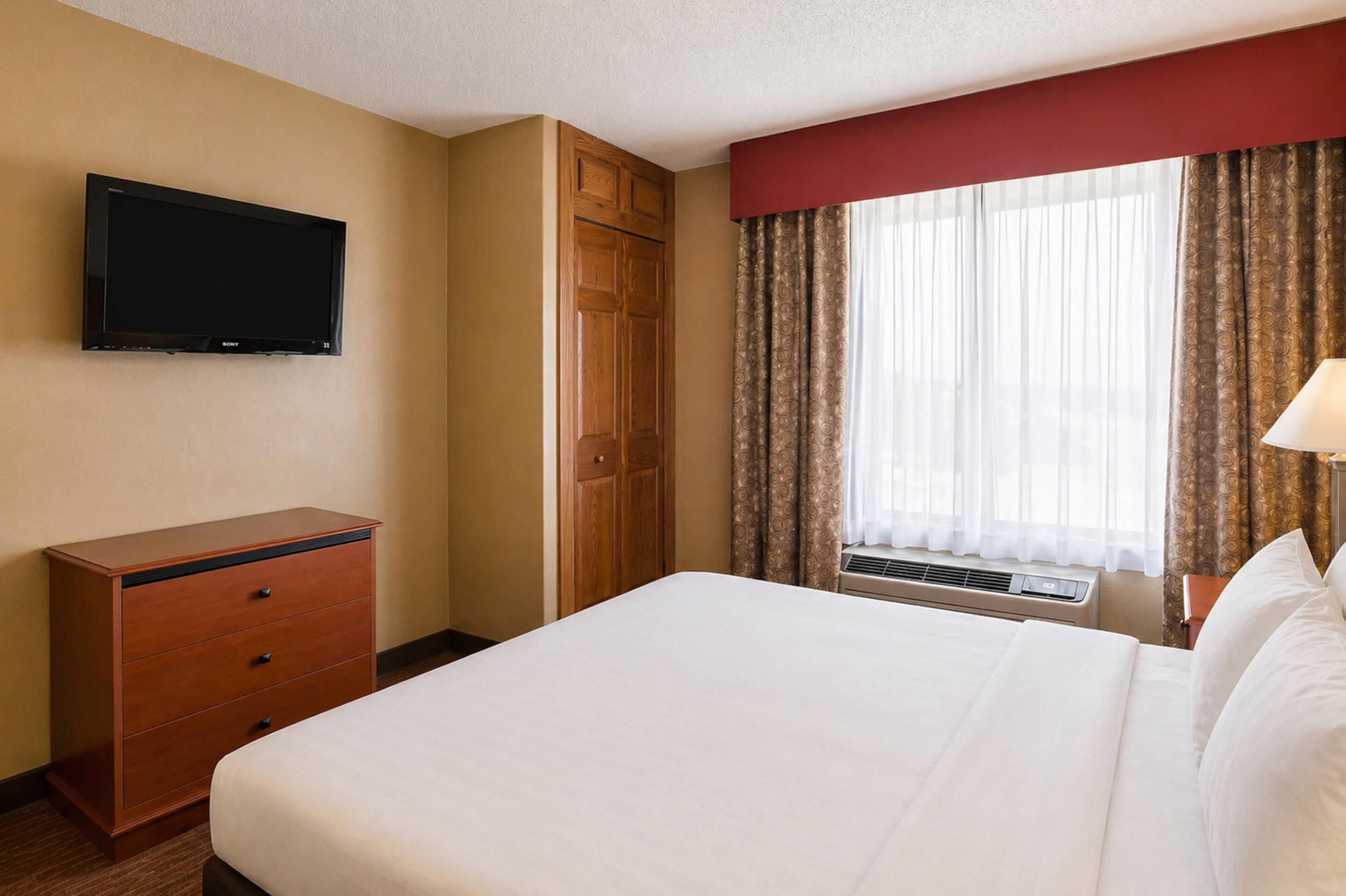 Quarto de hotel com cama, televisão fixada na parede, guarda-roupa de madeira e janela com cortinas.
