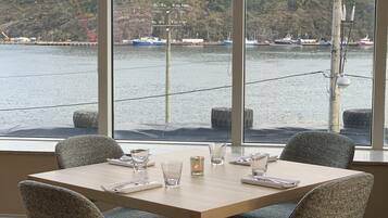 Uma sala de jantar com uma mesa posta para quatro pessoas, grandes janelas com vista para um porto com barcos e uma paisagem montanhosa.
