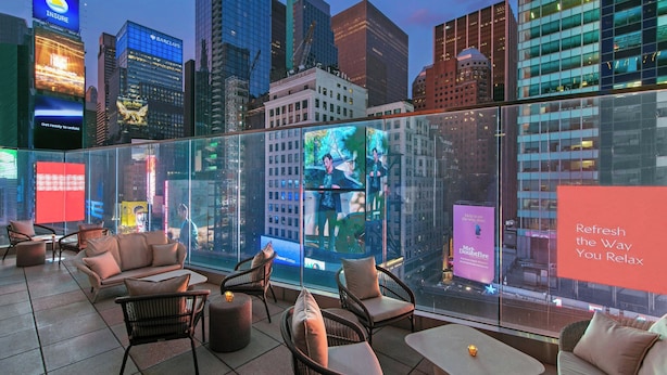 Um terraço com área de estar e móveis modernos, grandes janelas com vista para a cidade e telas digitais exibindo publicidade.
