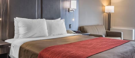 Quarto de hotel com uma cama grande, um cobertor vermelho, uma mesa de cabeceira com abajur e uma cadeira.