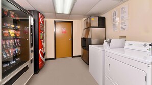 Uma lavanderia com máquina de lavar e secadora Whirlpool, uma máquina de vendas com lanches, e uma porta identificada como 'Sala de Equipamentos Elétricos'.