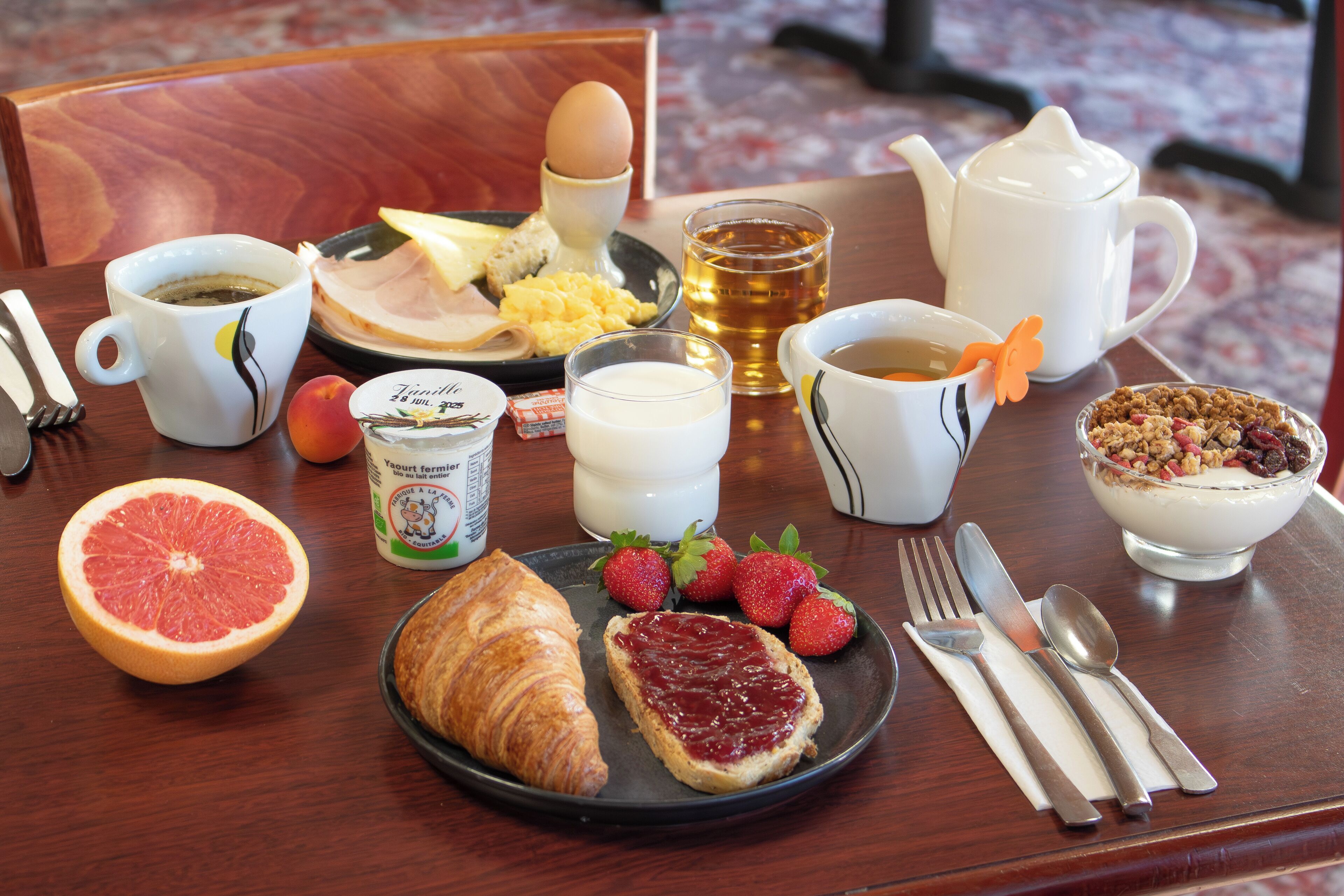 Uma mesa de café da manhã com croissants, morangos, uma toranja, um bule de chá, uma xícara de café, um copo de suco e uma tigela de cereais.