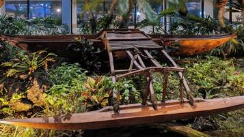 Um vaso em forma de barco com uma passarela de madeira, cercado por plantas tropicais e palmeiras.