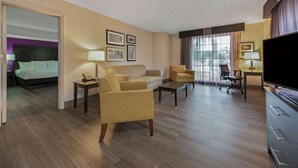 Quarto de hotel com cama, sofá, poltrona, mesa de centro, TV e escrivaninha com abajur.