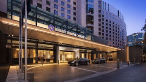 Entrada de um hotel moderno com fachada de vidro, rampa coberta e dois carros estacionados em frente.