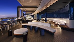Um moderno bar no terraço com área de estar curvada, piso de madeira e vista para a cidade ao entardecer.