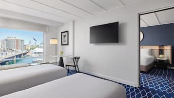 Quarto de hotel com duas camas, televisão, escrivaninha com cadeira, mesa pequena com abajur e vista para a paisagem urbana e para a água.