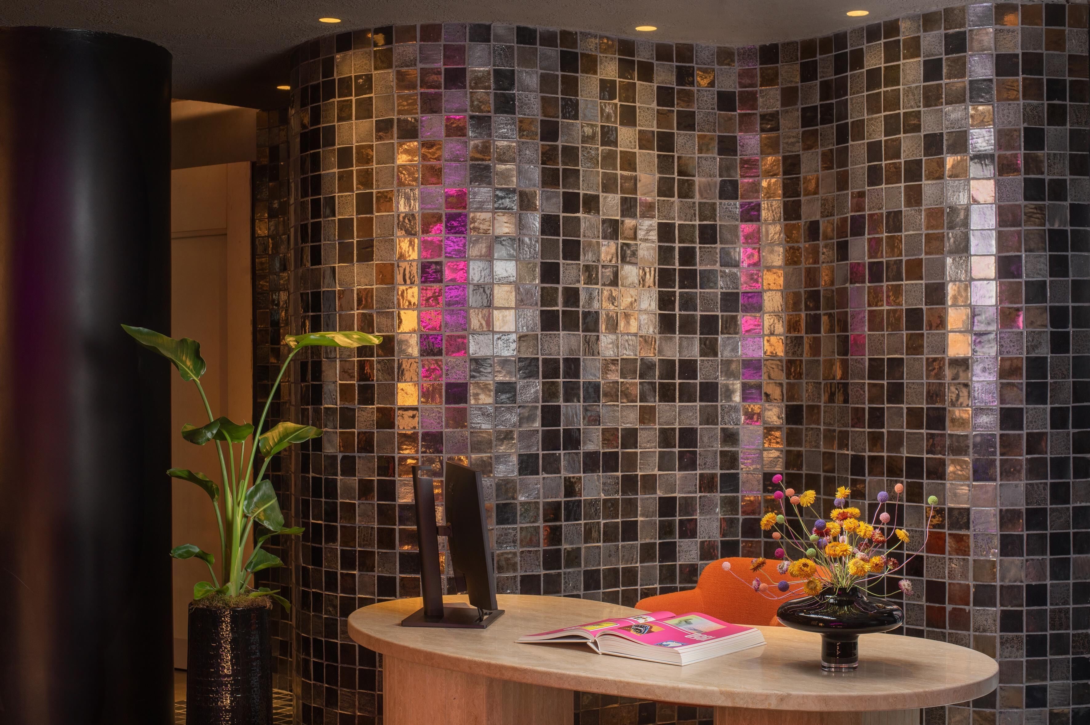 Uma área de recepção com um painel curvo de mosaicos, balcão de atendimento, planta em vaso e um arranjo de flores.