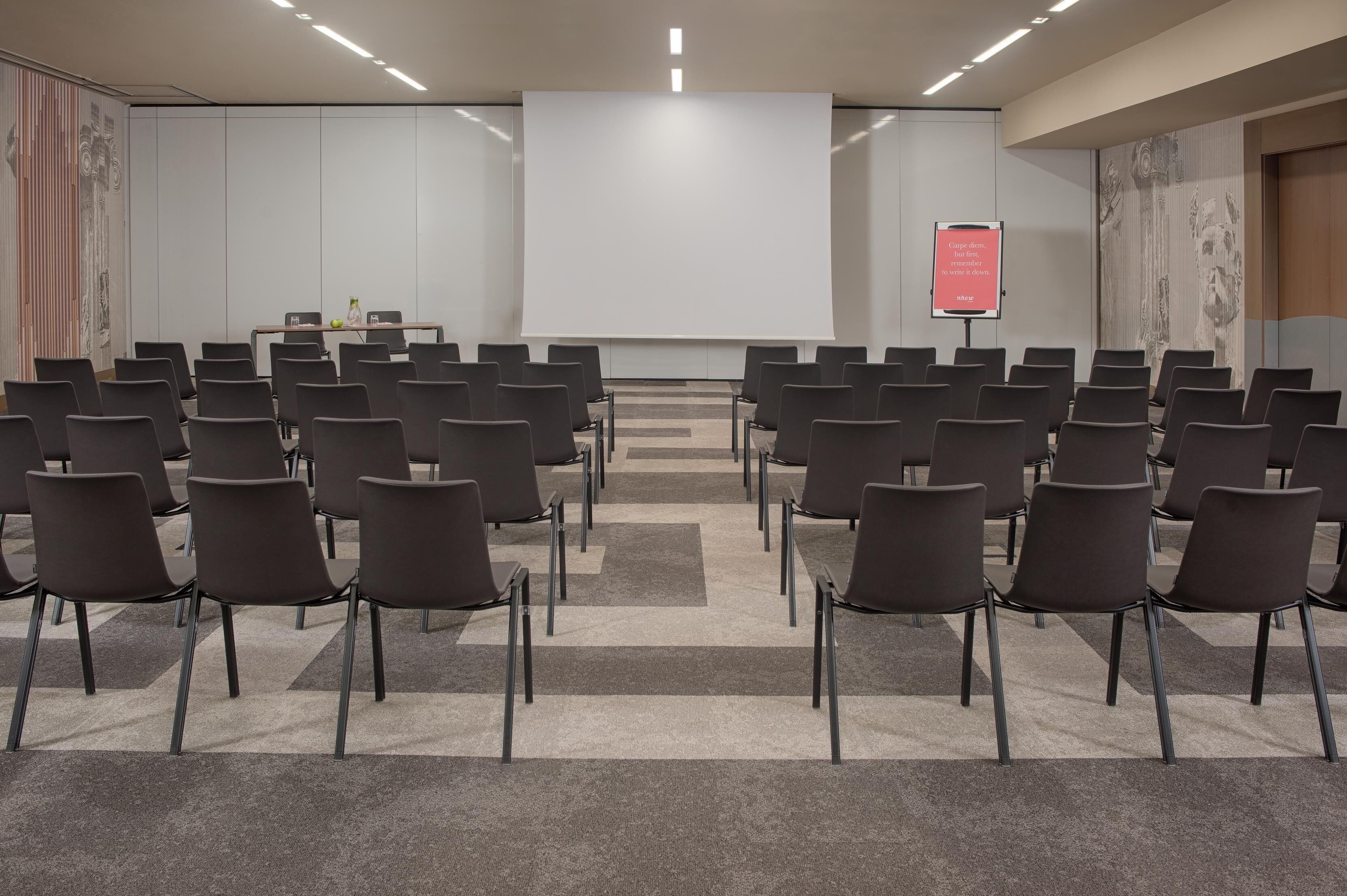 Uma sala de conferências com fileiras de cadeiras voltadas para uma tela grande, um púlpito e um quadro branco.