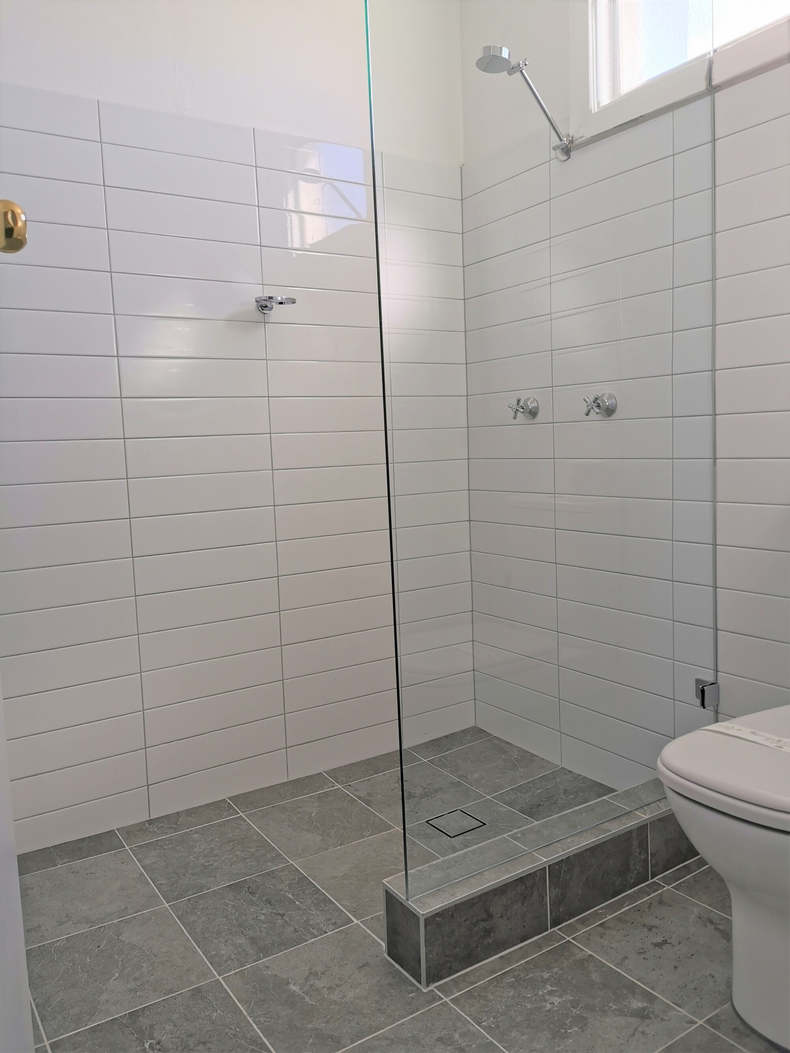 Casa de banho moderna com paredes revestidas a azulejos brancos, cabine de duche em vidro, chão em mosaico cinzento e sanita branca.