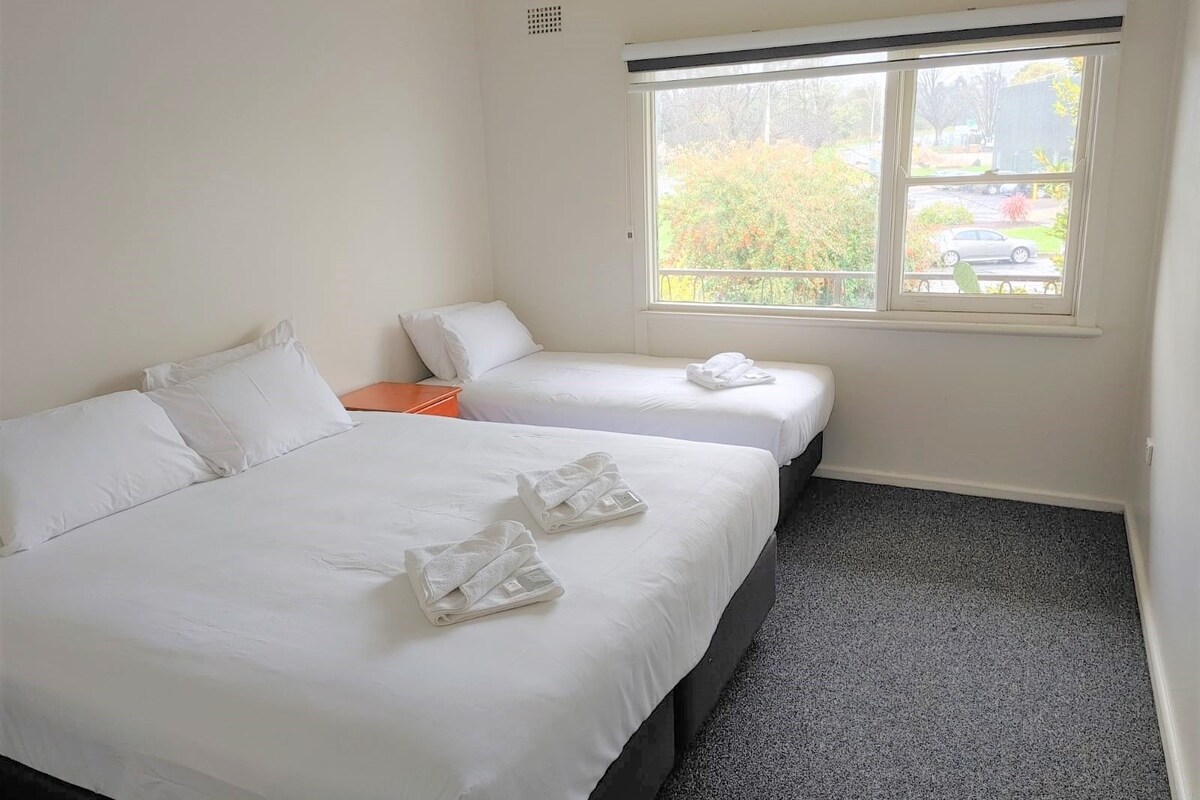 Quarto de hotel com duas camas, janela com vista para árvores e estacionamento, e piso coberto de carpete.