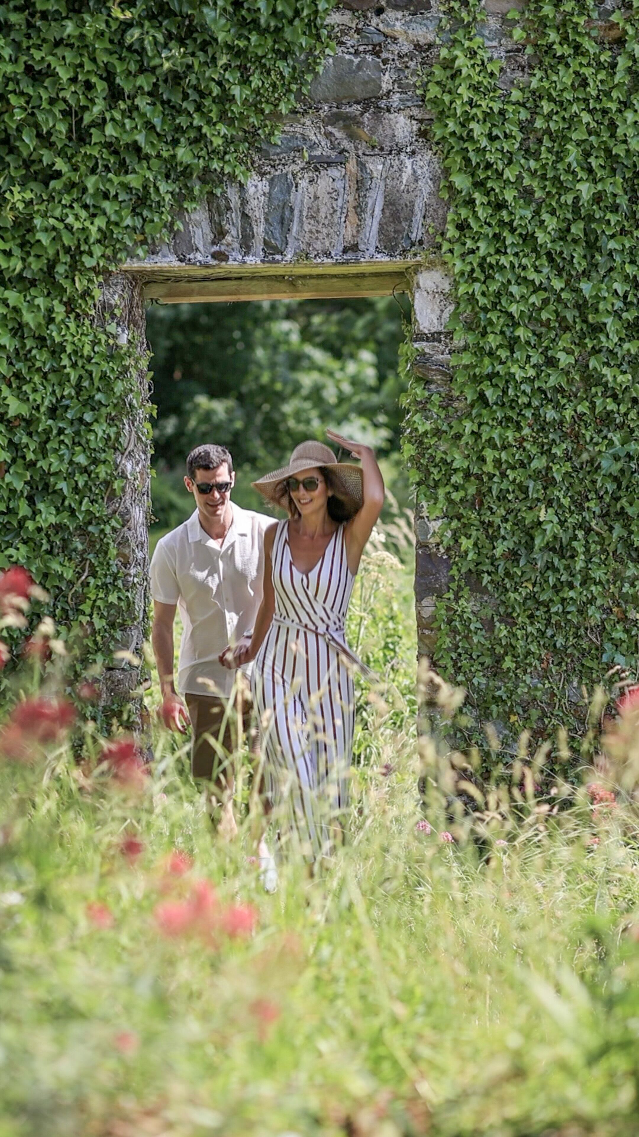 Um casal caminhando por um jardim com um arco de pedra coberto de hera.