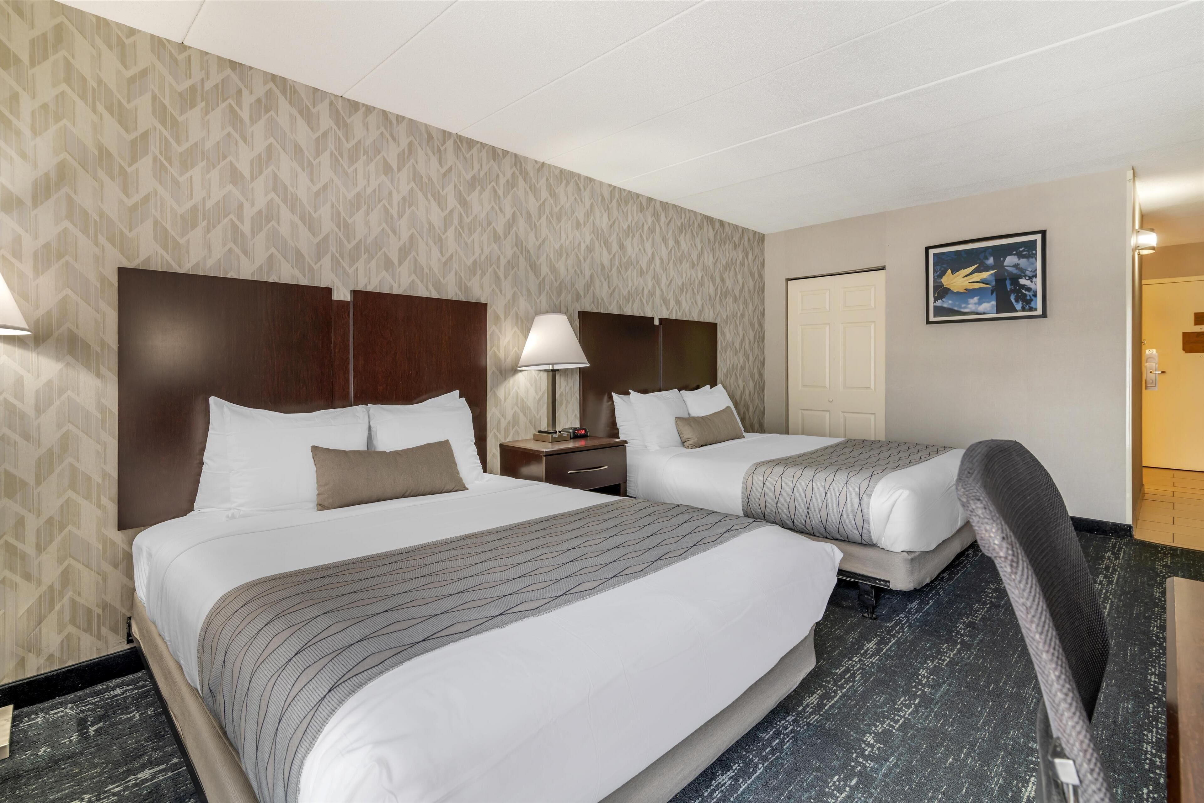Quarto de hotel com duas camas, uma mesa de cabeceira com abajur, uma cadeira e uma porta.