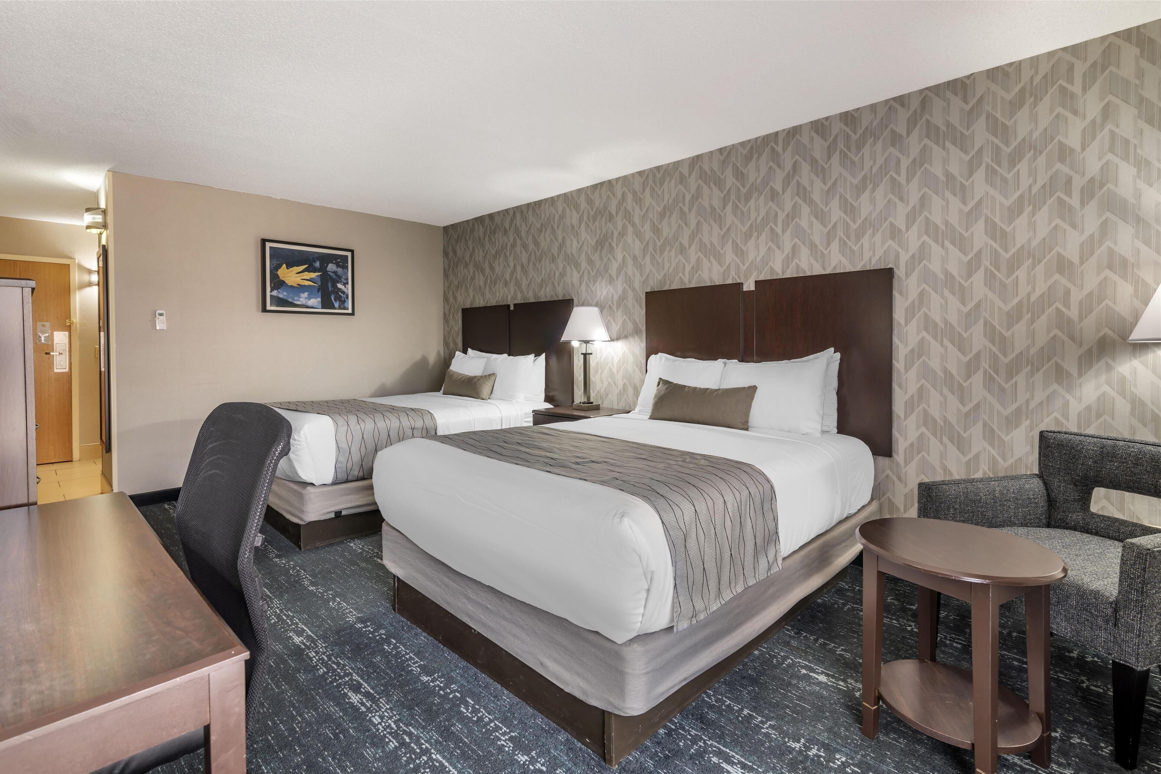 Quarto de hotel com duas camas, uma escrivaninha, uma cadeira e uma mesinha.