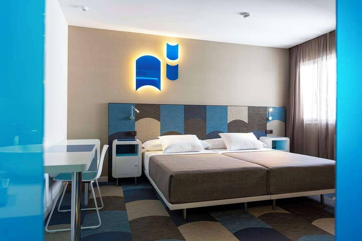Quarto de hotel com cama, mesas de cabeceira, escrivaninha e cadeiras.