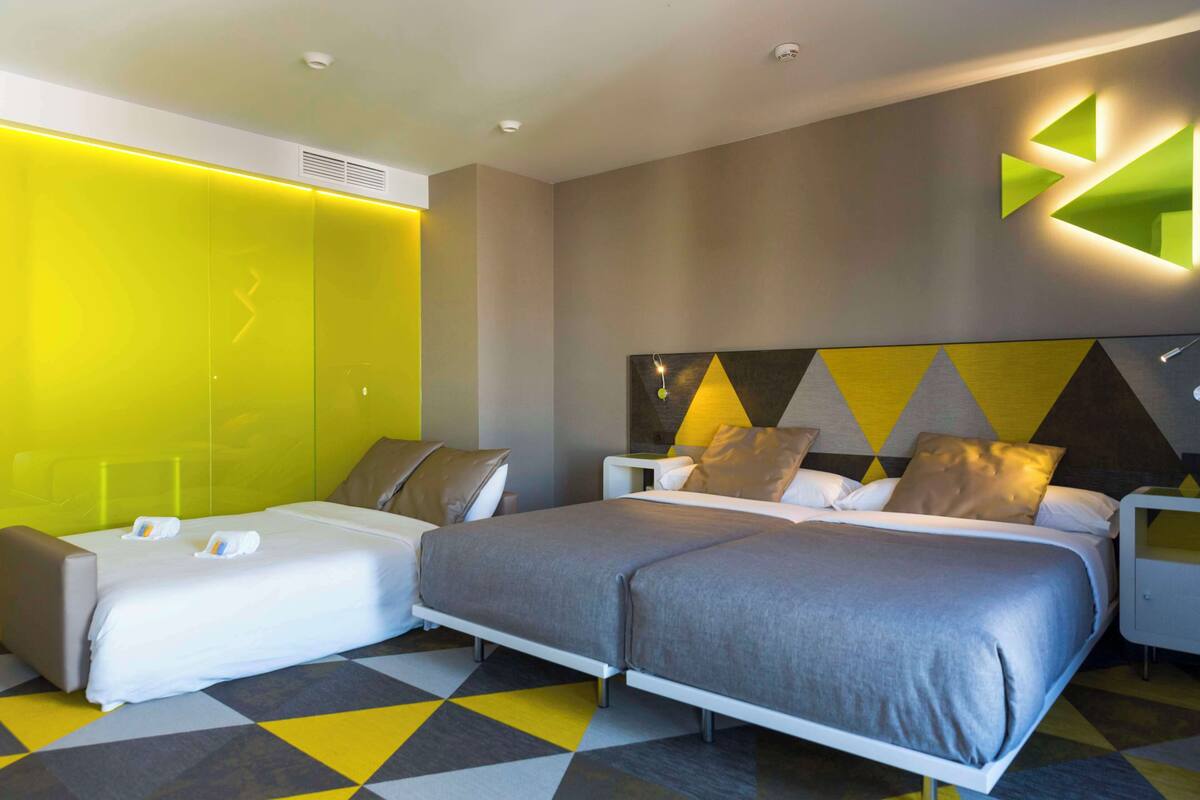 Quarto de hotel moderno com duas camas, um sofá e um chão com padrão geométrico.