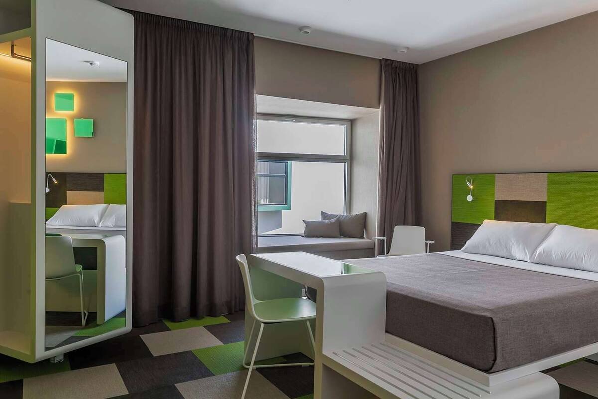Quarto de hotel moderno com cama, escrivaninha e cadeira. Possui uma janela com cortinas, e o quarto segue um esquema de cores verde e bege.