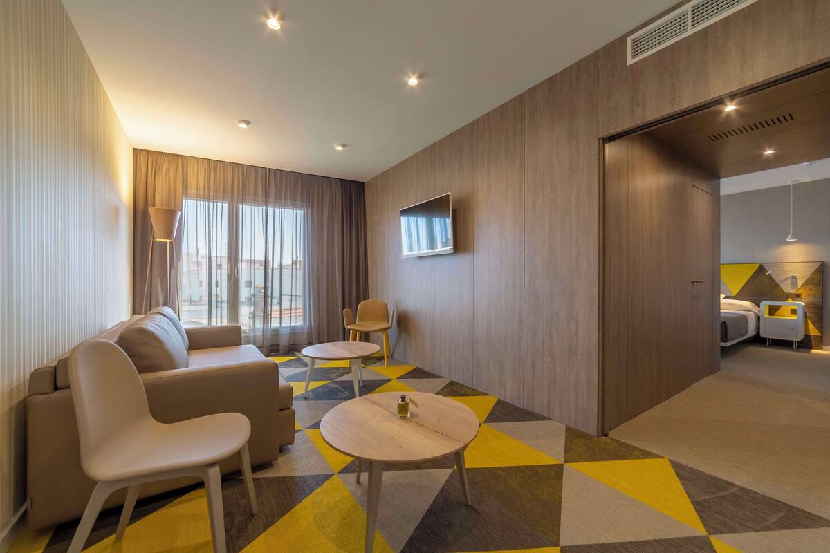 Quarto de hotel moderno com sofá, duas cadeiras, uma mesinha e televisão fixada na parede.