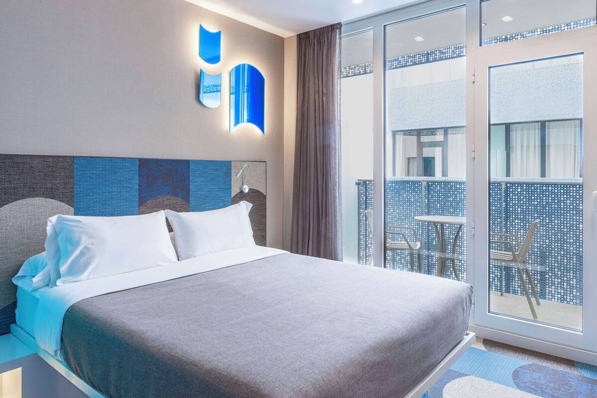 Quarto de hotel moderno com uma cama grande, porta de vidro que dá para uma varanda com mobiliário de exterior e vista para a cidade.