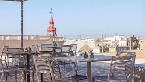 Área externa com mesas e cadeiras, com vista para um horizonte urbano onde se destaca uma torre vermelha.
