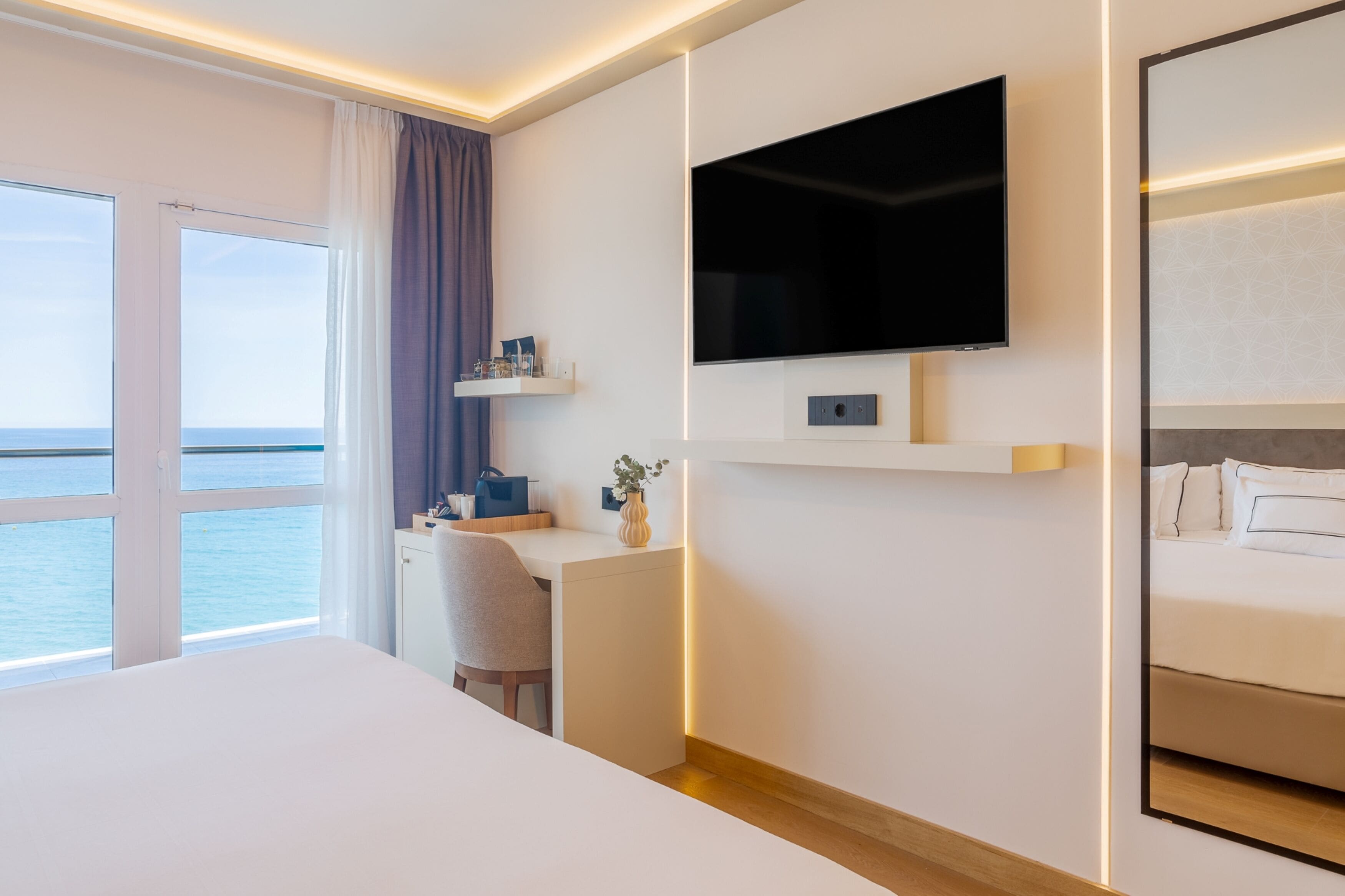 Quarto de hotel moderno com uma cama grande, uma escrivaninha com cadeira, televisão na parede e vista para o mar pela janela.