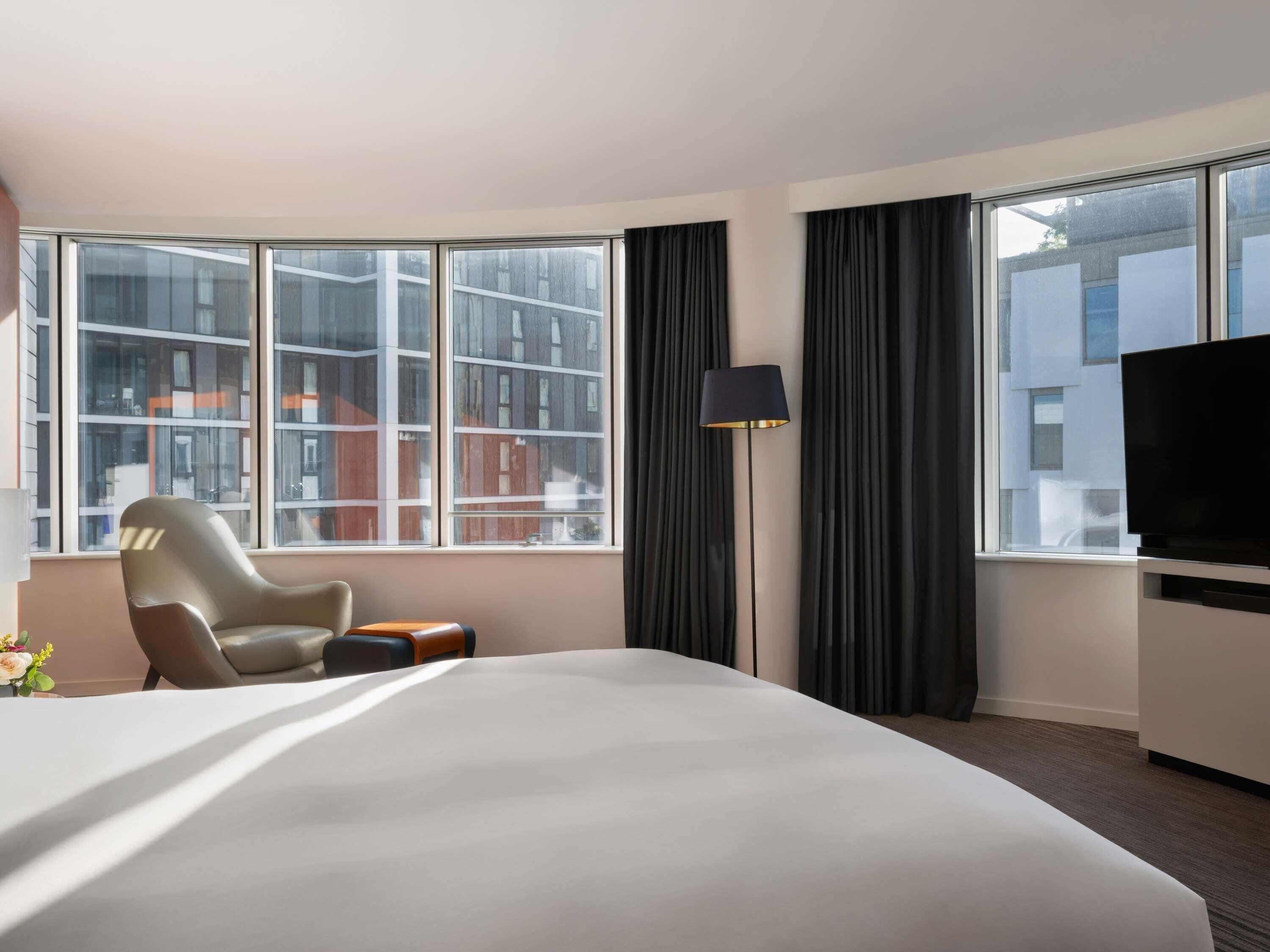 Quarto de hotel com uma cama grande, uma cadeira, uma televisão, um abajur e uma janela com cortinas.