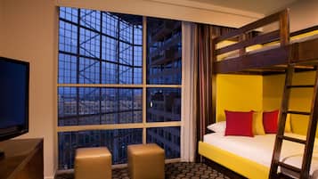 Quarto de hotel com cama de solteiro, televisão, janela ampla com vista para a cidade e dois pufes.