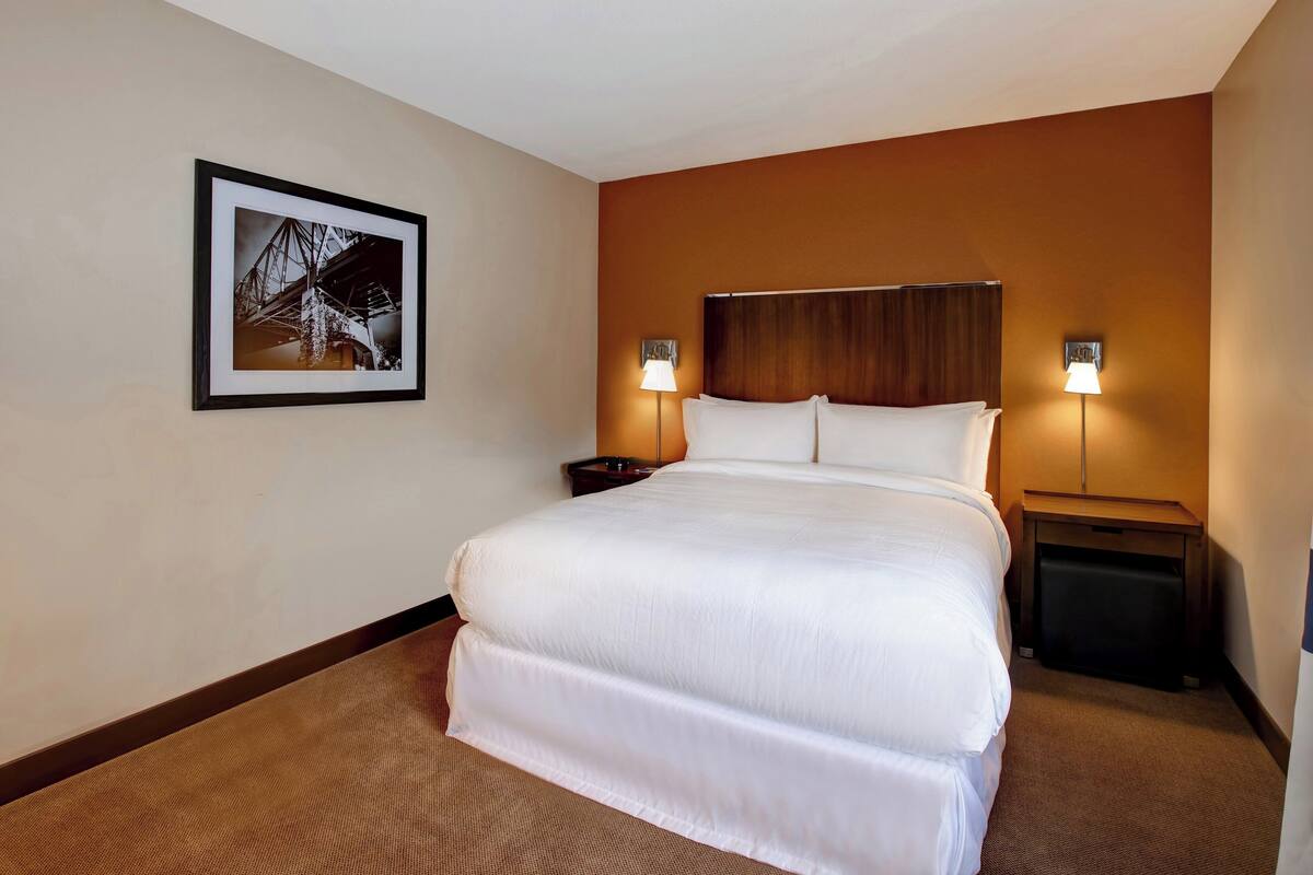 Quarto de hotel com uma cama grande, duas luminárias de cabeceira, um quadro na parede e uma pequena mesa de apoio.