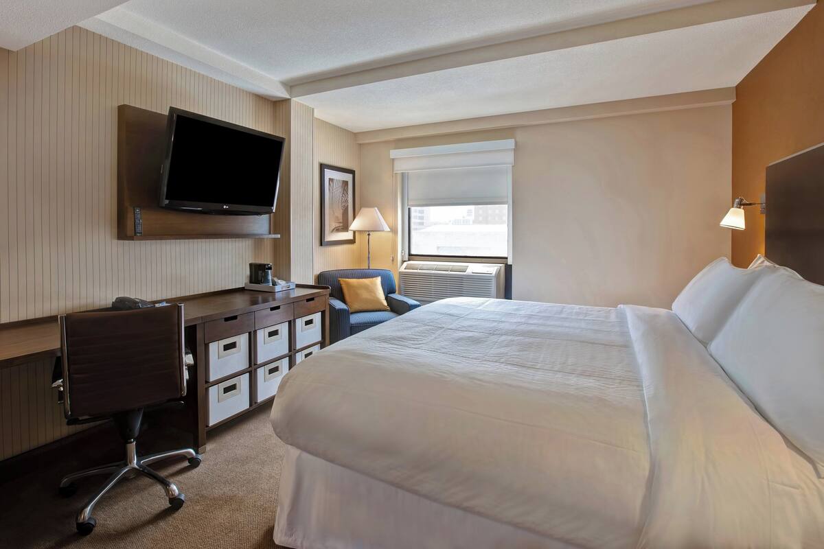 Quarto de hotel com uma cama grande, uma escrivaninha com cadeira, televisão na parede, abajur e uma janela com persianas.