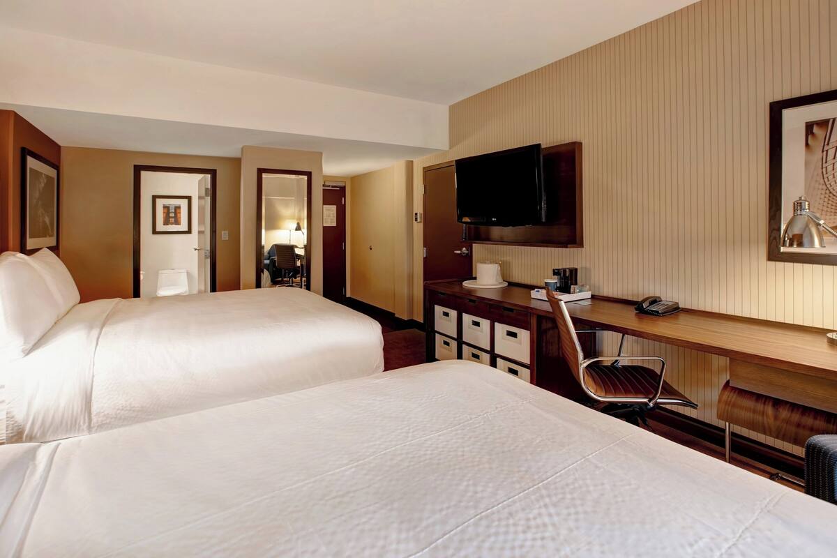 Quarto de hotel com duas camas, uma mesa de trabalho com cadeira, televisão e espelho.