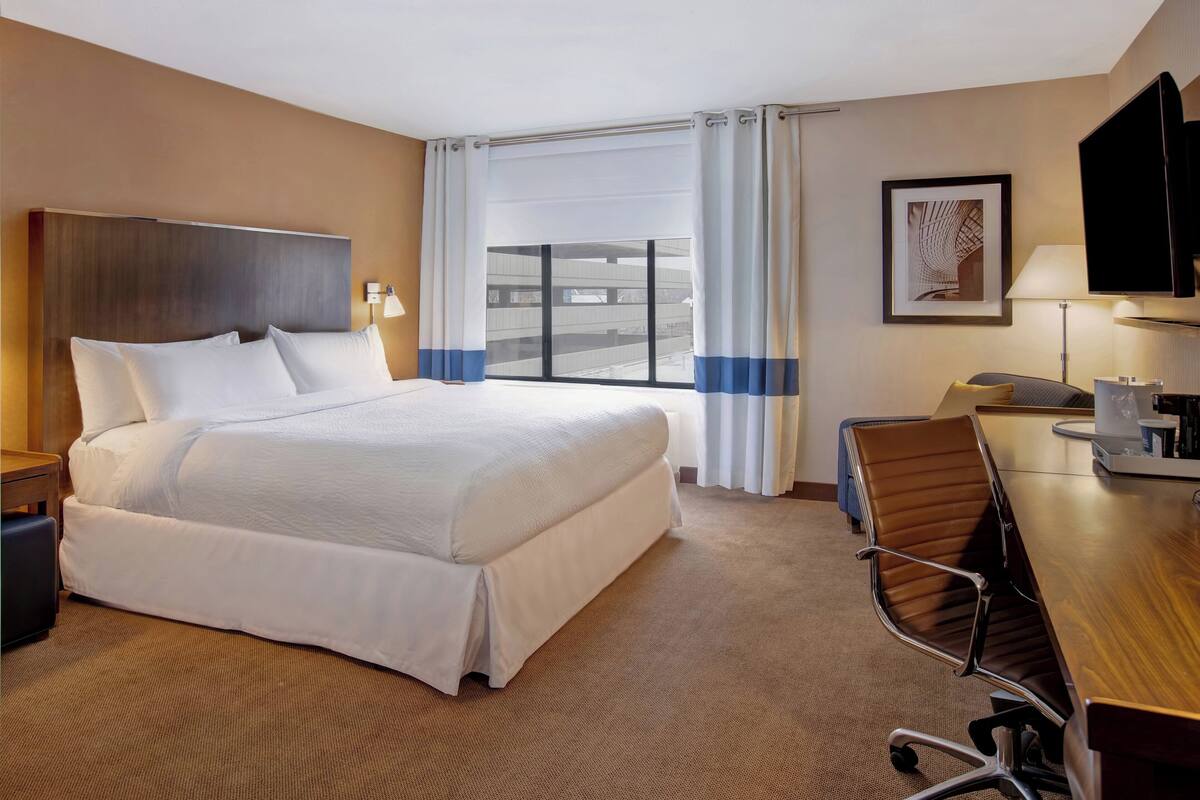 Quarto de hotel com uma cama grande, uma escrivaninha com cadeira, televisão de tela plana e janela com cortinas.