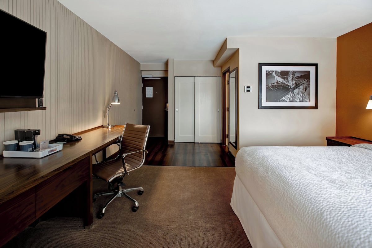 Quarto de hotel com cama, escrivaninha, cadeira, televisão, candeeiro e um quadro na parede.