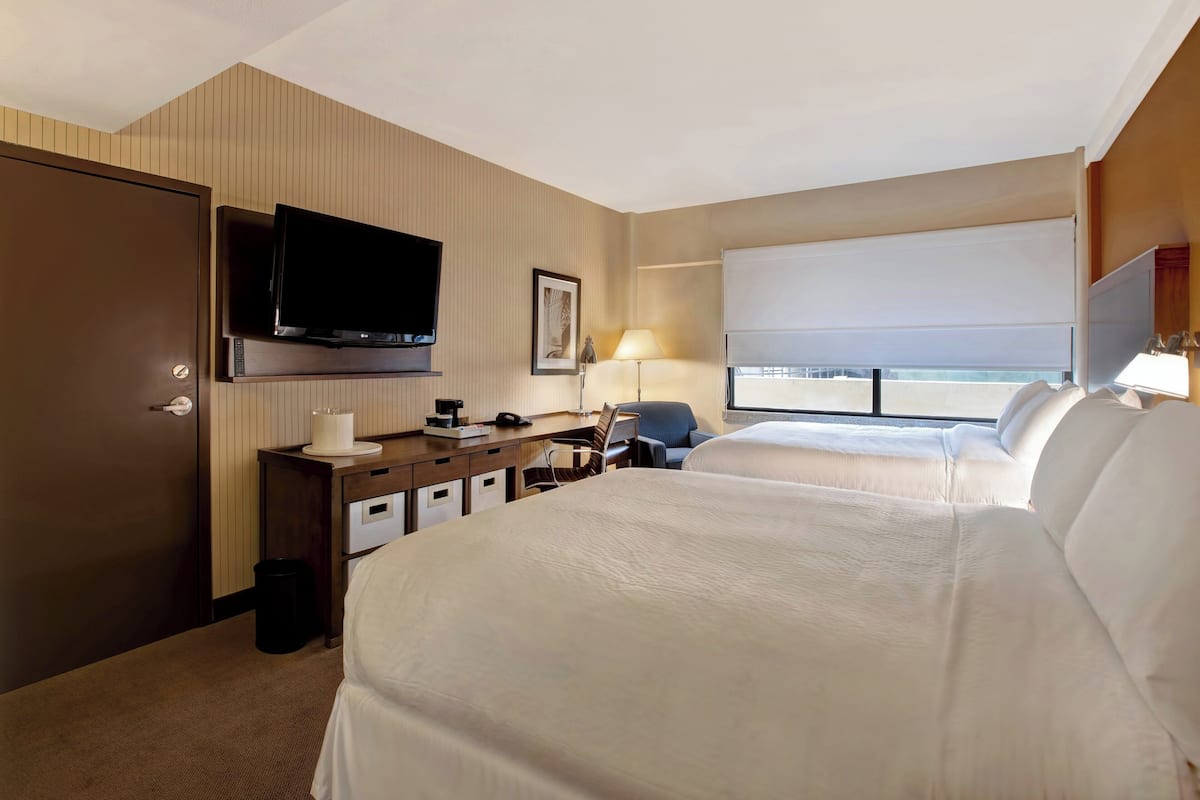 Quarto de hotel com duas camas, uma TV, uma escrivaninha com abajur e uma janela com vista.